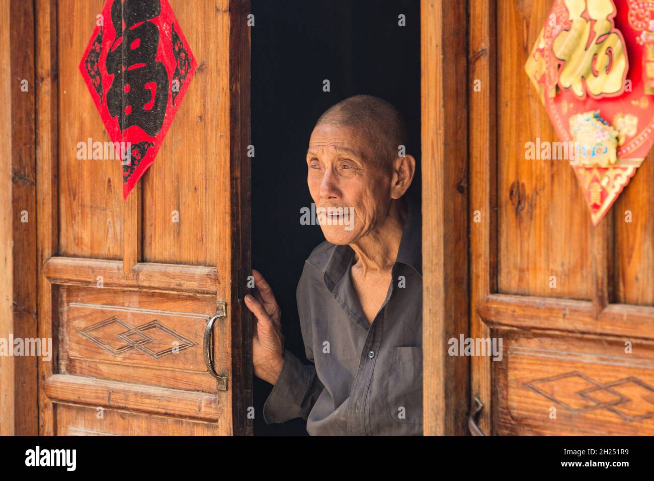 An elderly Chinese man looks out the doorway of his traditional house ...