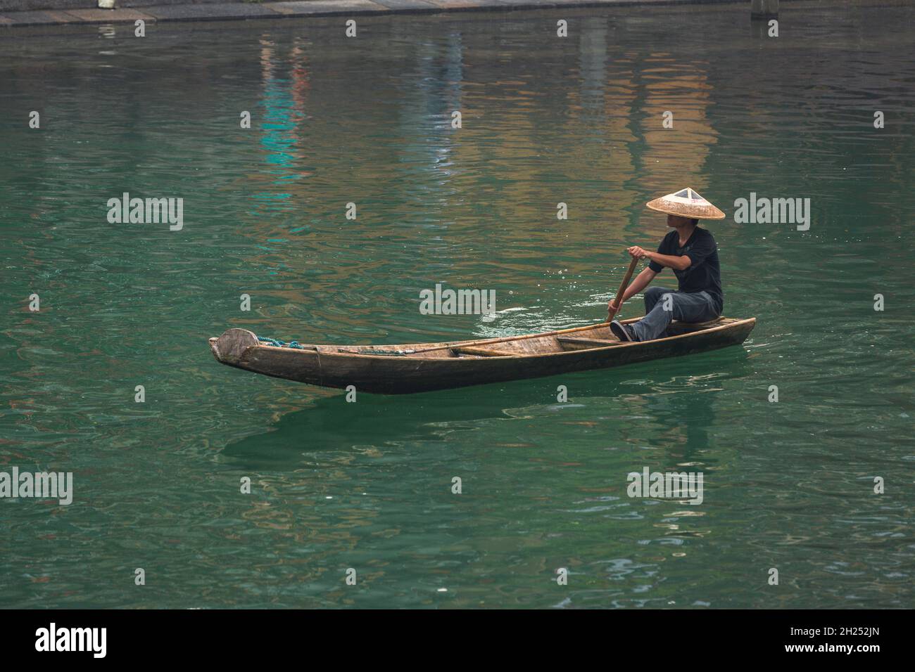 A man in a traditional conical hat paddles a sampan on the Tuojiang ...