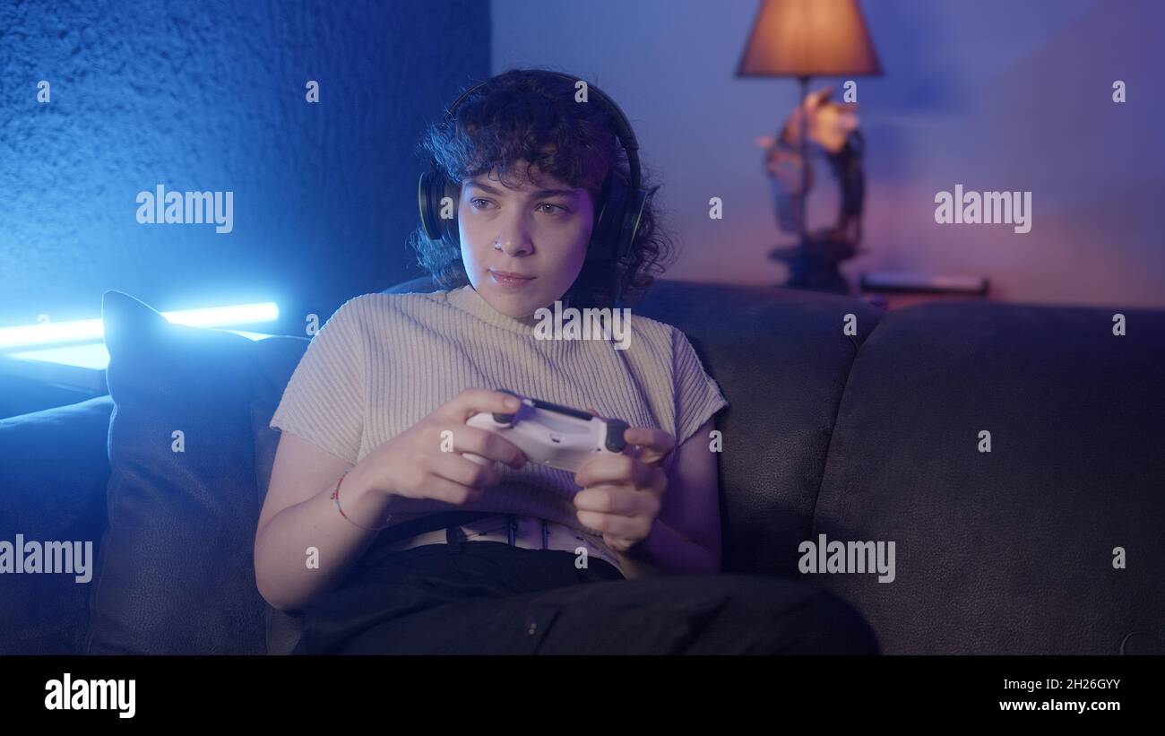 Young curly haired gamer woman with headphone sitting on sofa relaxing ...