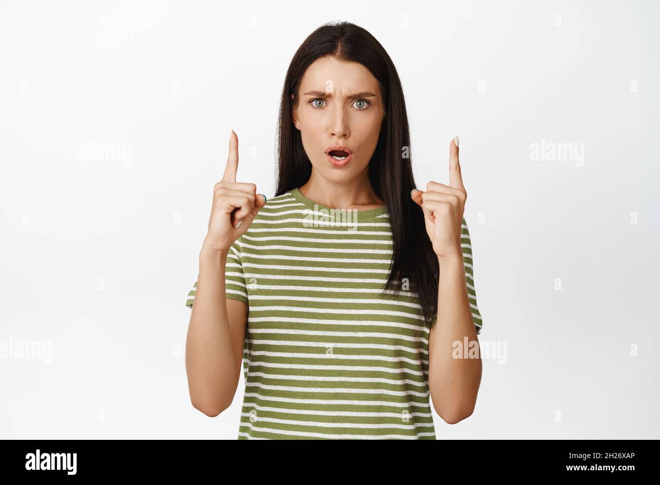 Confused brunette woman pointing fingers up, looking puzzled and ...