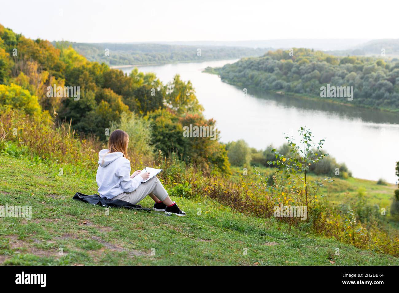 A student girl is sitting on the river bank and writing in a notebook ...