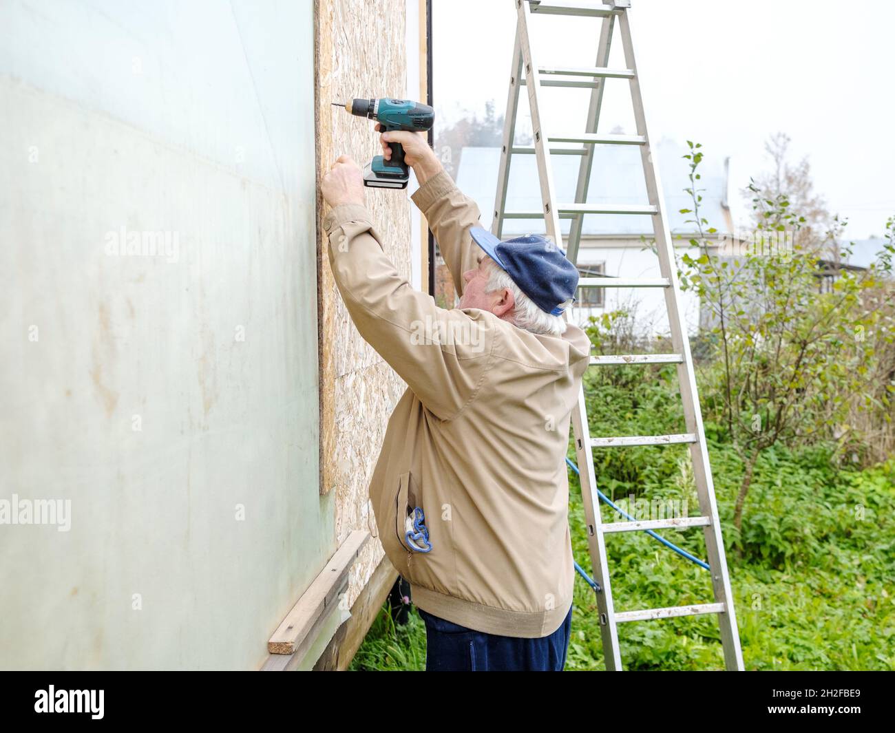 An elderly gray-haired carpenter screws an oriented strand board to the ...