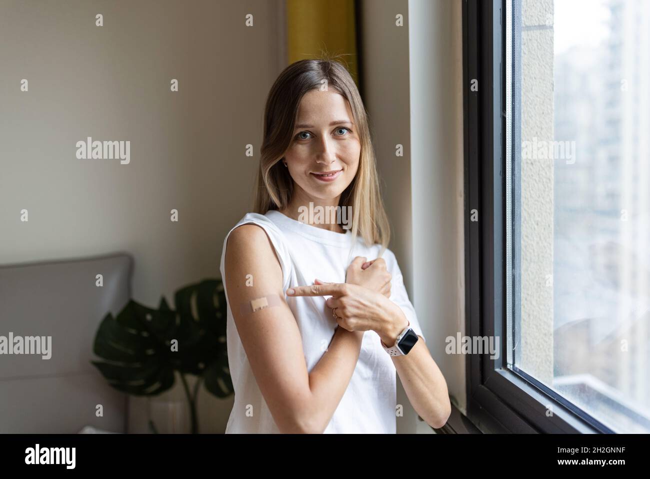 Happy young caucasian woman with blonde hair in white t-shirt showing patch on arm, feeling good ...