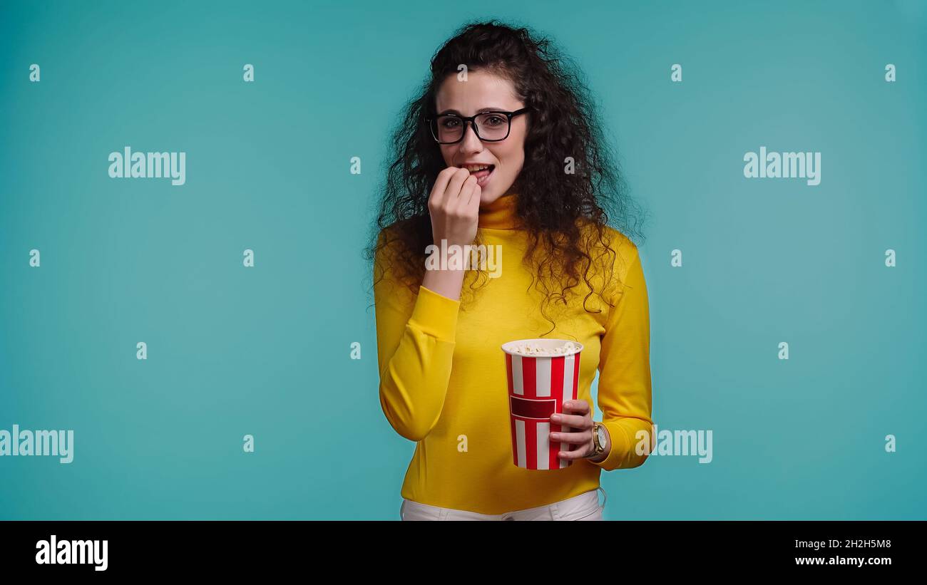 curly young woman eating popcorn while watching movie isolated on blue ...