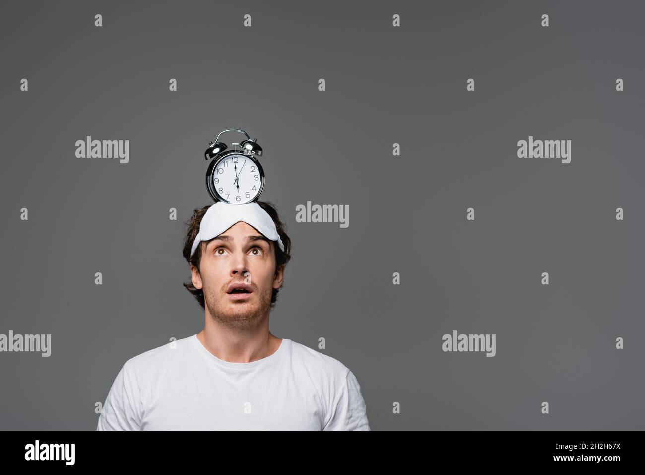 Shocked man in sleep mask looking at alarm clock on head isolated on ...