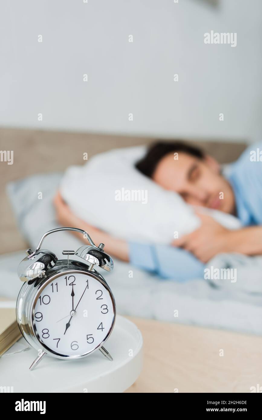 Alarm clock and book on bedside table near blurred man sleeping on bed ...