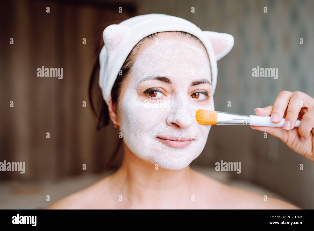 Woman with a white clay mask on her face holds a brush. Female takes ...