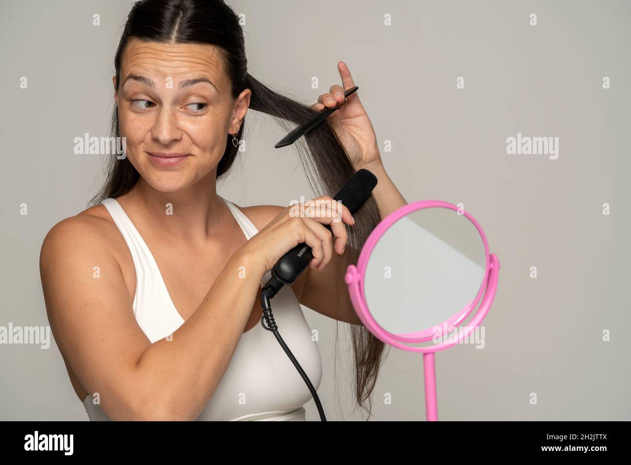 Young smiling woman straightening her hair with a straightener on gray ...
