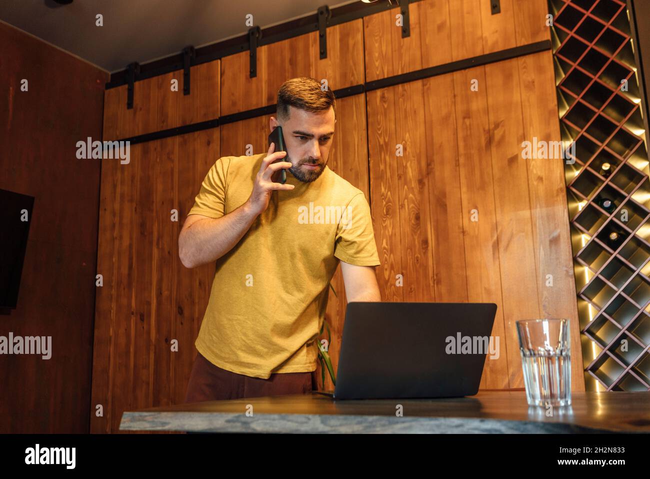 Businessman talking on smart phone while using laptop in living room ...