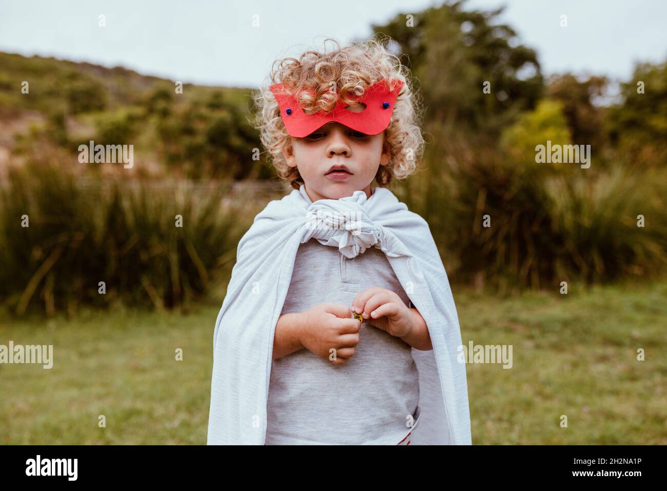 Cute blond boy with curly hair wearing mask and cape standing on meadow ...