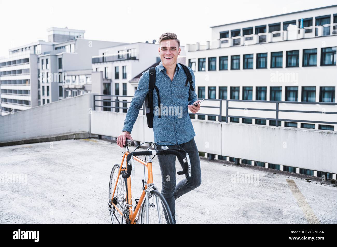 Smiling man with smart phone wheeling bicycle while walking on rooftop ...