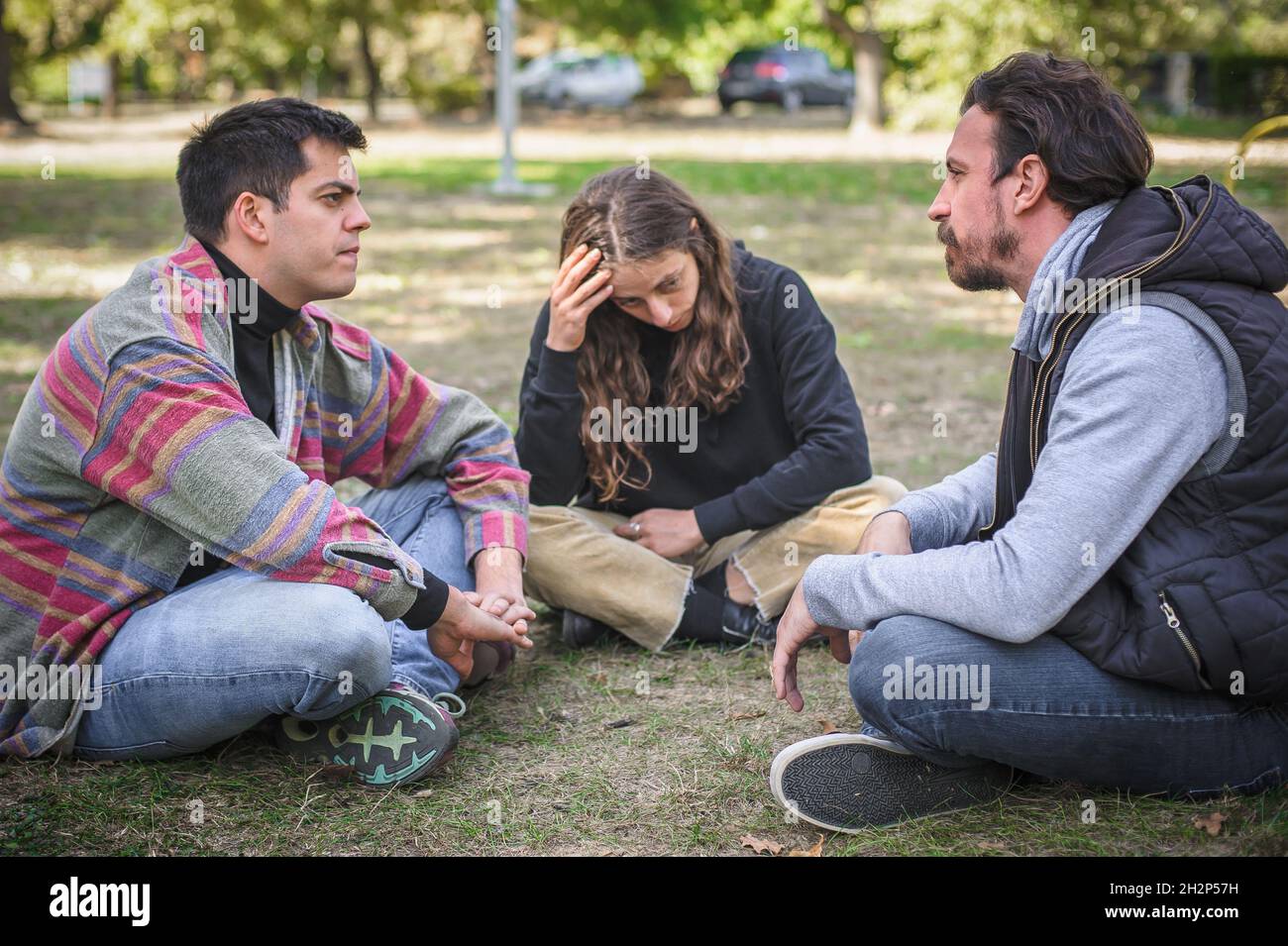 Three worried friends, two men and a girl, sit together on the ground ...