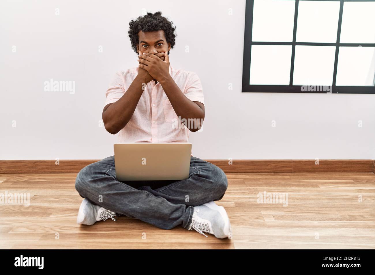 African man with curly hair using laptop sitting on the floor shocked ...