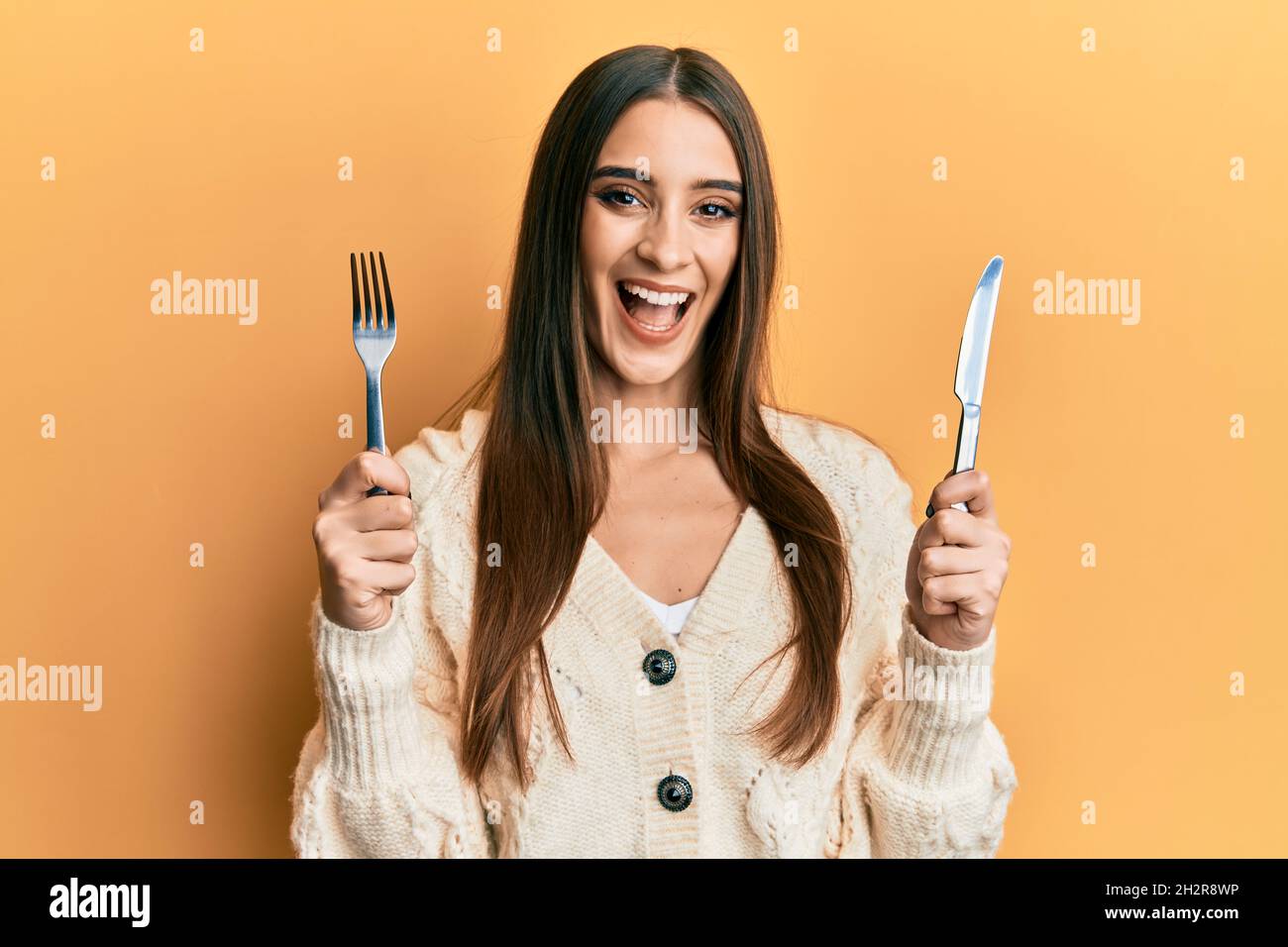 Beautiful brunette young woman holding fork and knife ready to eat smiling and laughing hard out ...