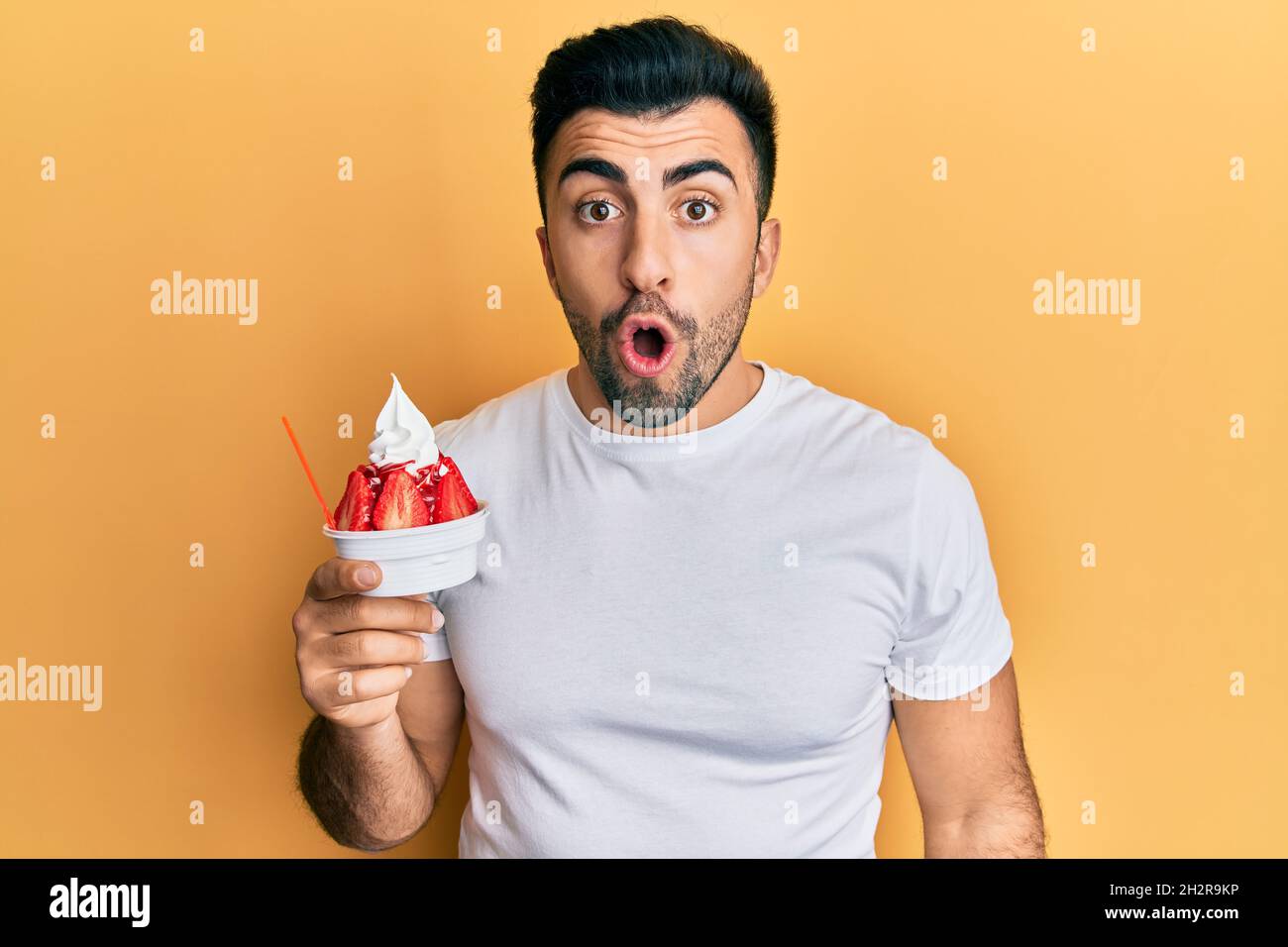Young hispanic man eating strawberry ice cream scared and amazed with ...
