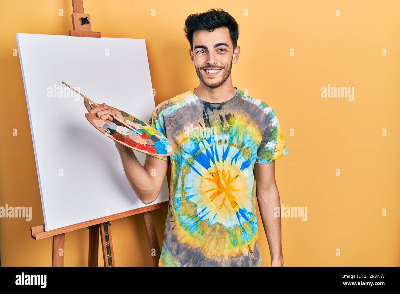 Young hispanic man standing drawing with palette by painter easel stand ...