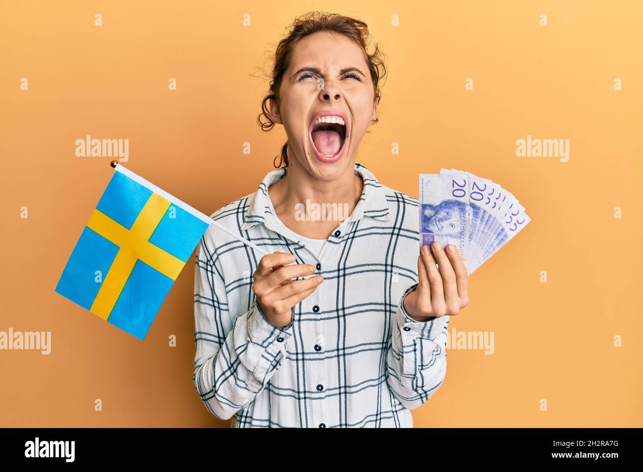 Young brunette woman holding sweden flag and krone banknotes angry and ...