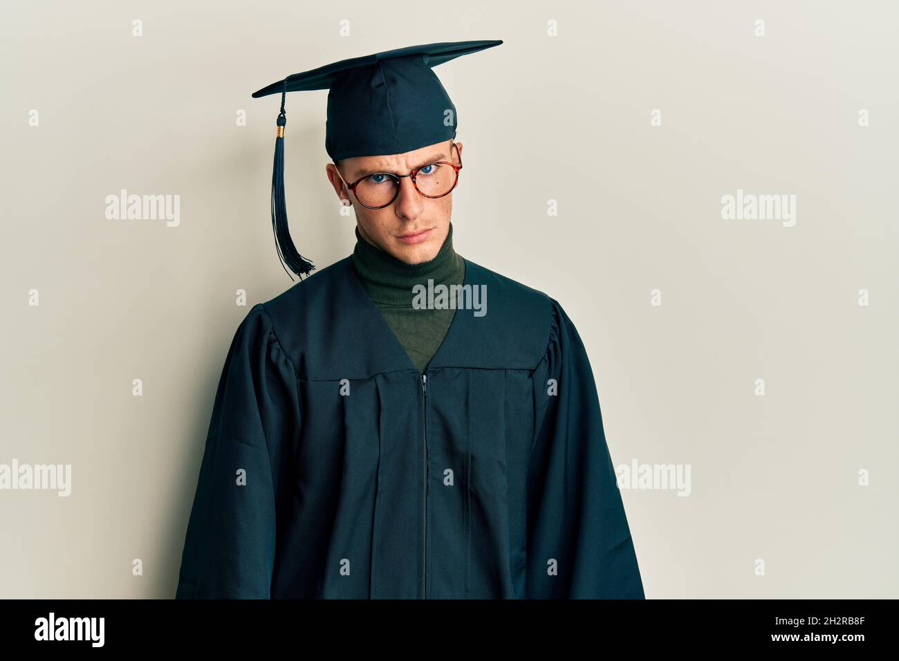 Young caucasian man wearing graduation cap and ceremony robe skeptic ...