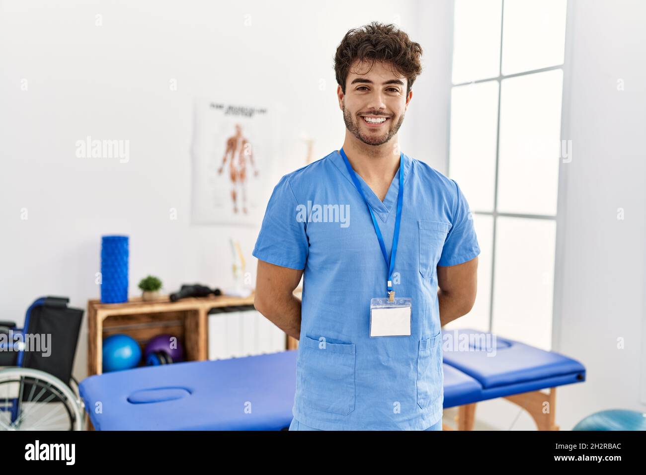 Young hispanic man wearing physio therapist uniform standing at clinic ...