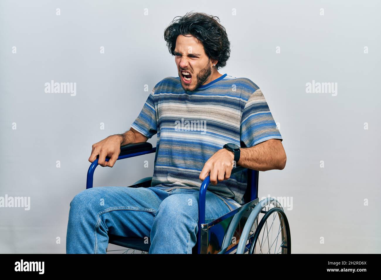 Handsome hispanic man sitting on wheelchair angry and mad screaming ...