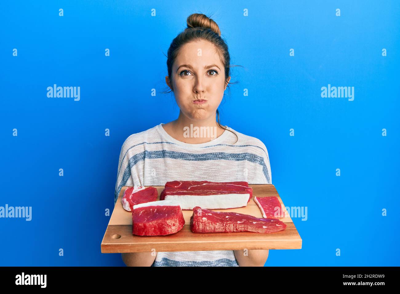 Young blonde woman holding board with raw meat puffing cheeks with ...