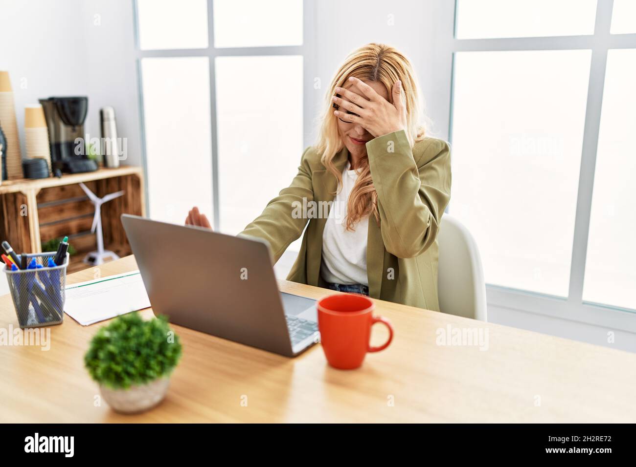 Beautiful blonde woman working at the office with laptop covering eyes ...