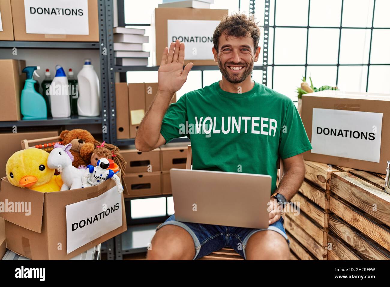 Young handsome man wearing volunteer t shirt using laptop waiving ...