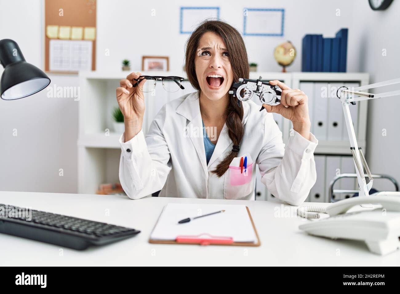 Young doctor woman holding optometry glasses and normal glasses angry ...