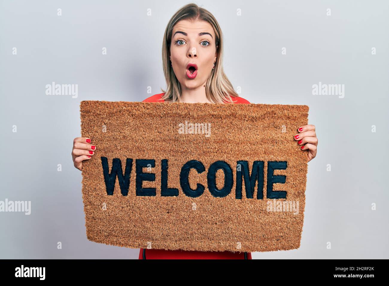 Beautiful caucasian woman holding welcome doormat afraid and shocked ...