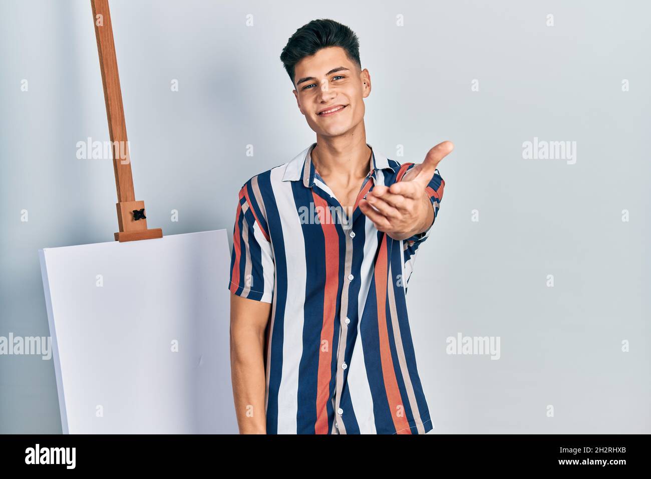Young hispanic man standing close to empty canvas smiling friendly ...