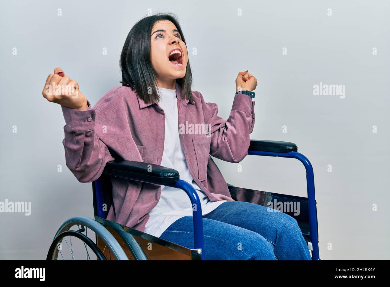 Young brunette woman sitting on wheelchair crazy and mad shouting and ...