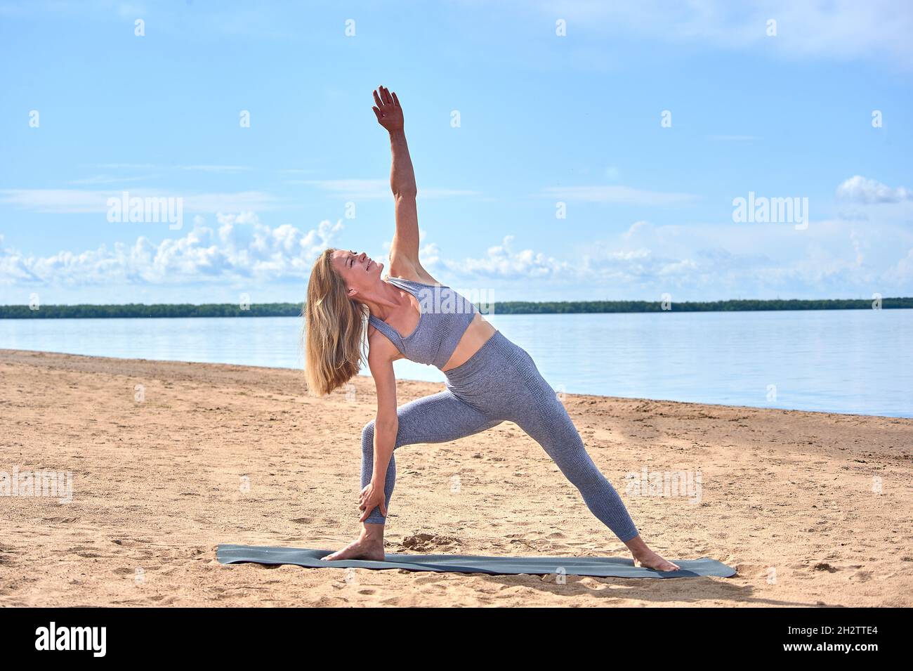 A woman does yoga on a sandy beach against the background of a river ...
