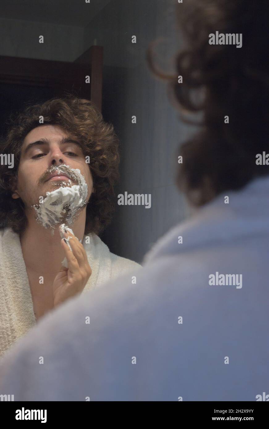 Young man in bathrobe applies shaving foam on face in apartment bathroom. Beauty Home Spa ...