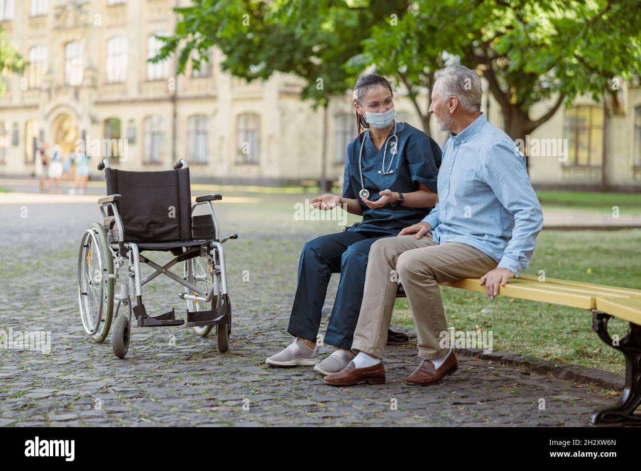 Talkative caring young nurse wearing face shield chatting with aged man ...