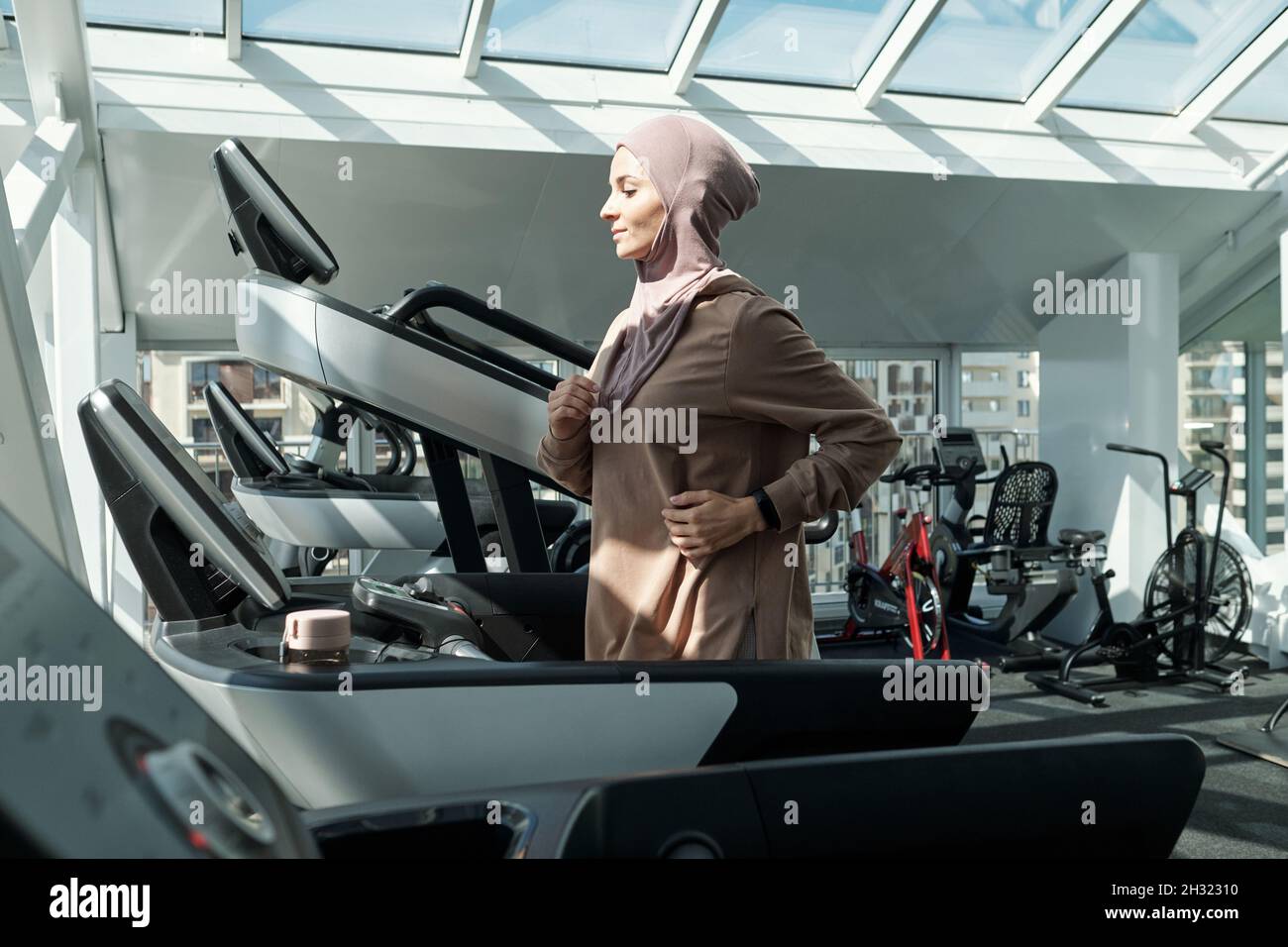 Young Muslim female in hijab and sportswear running on treadmill during ...