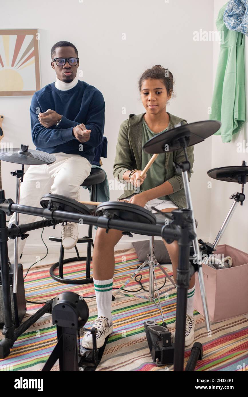 Cute schoolgirl hitting cymbal of drumkit during home lesson or ...