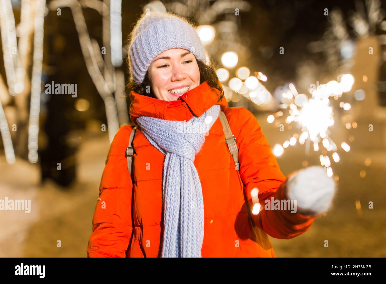 Smiling young woman wearing winter knitted clothes holding sparkler outdoors over snow ...