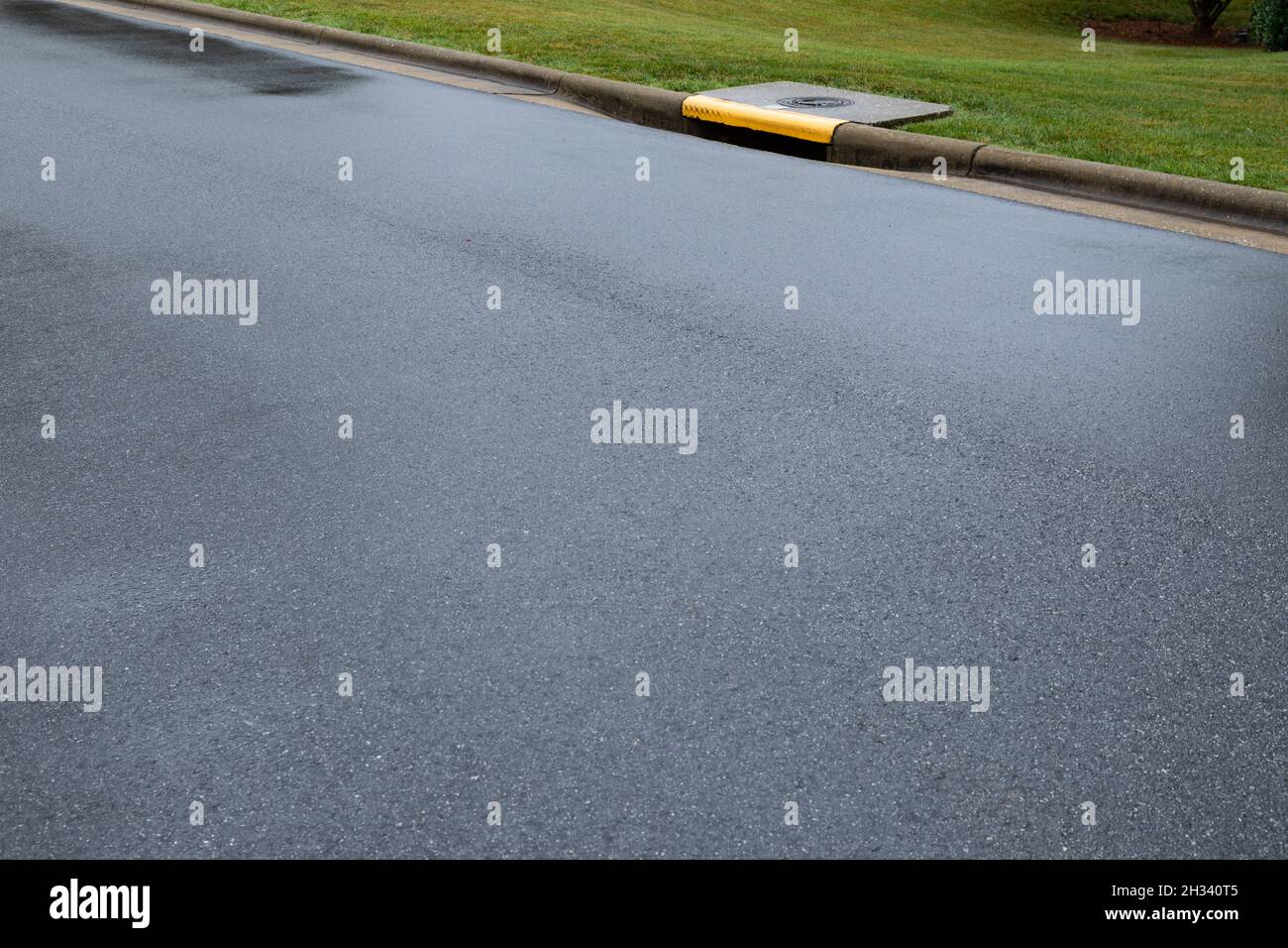 Dark and wet asphalt road bed with curb, grass, and storm drain, ample ...