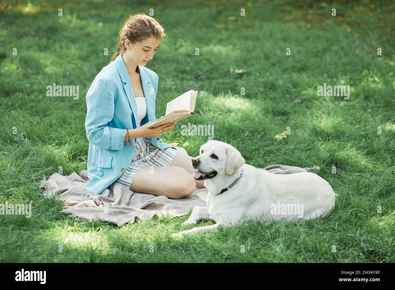 Full length portrait of young woman reading book outdoors with Labrador ...