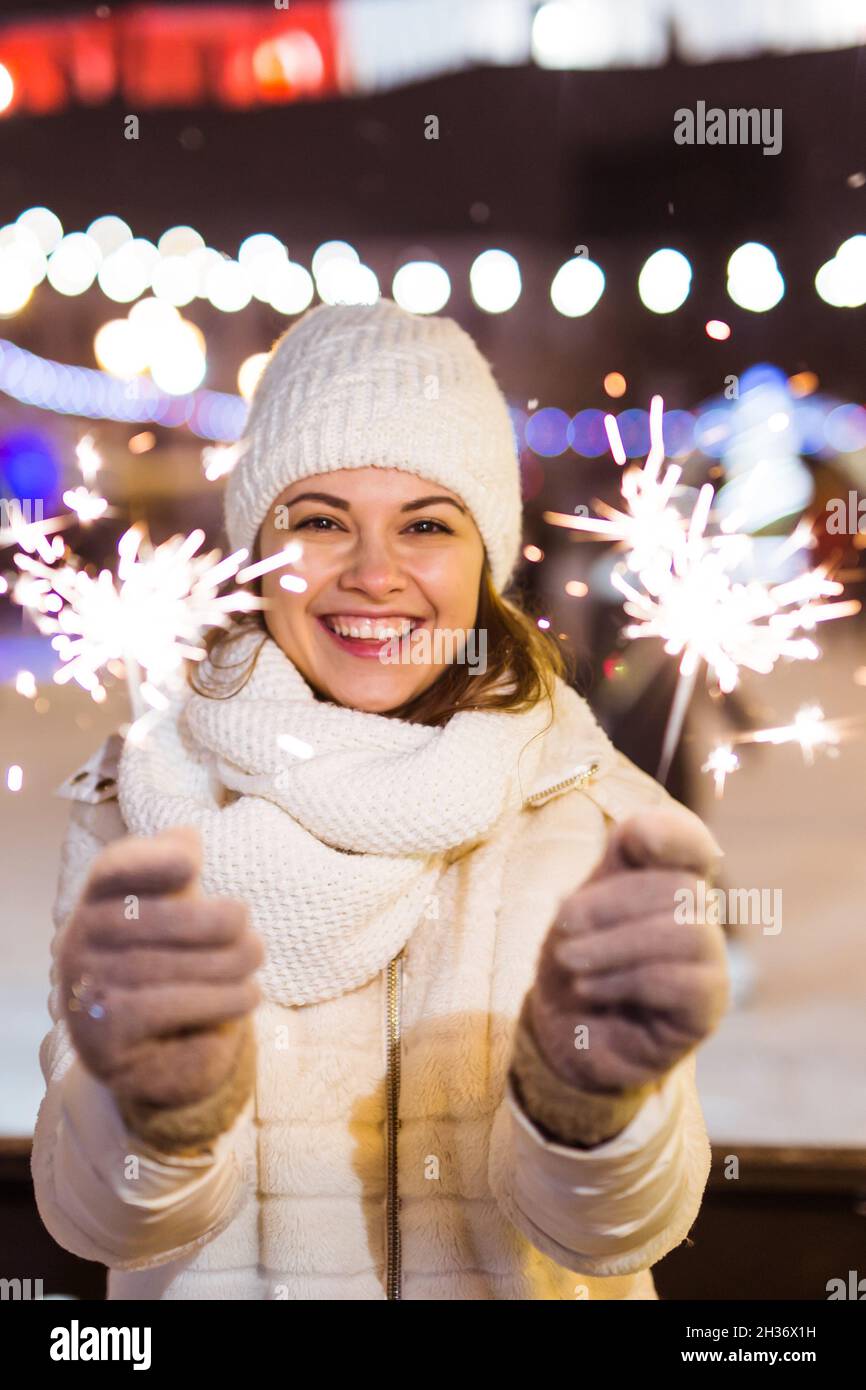 Smiling young woman wearing winter knitted clothes holding sparkler outdoors over snow ...