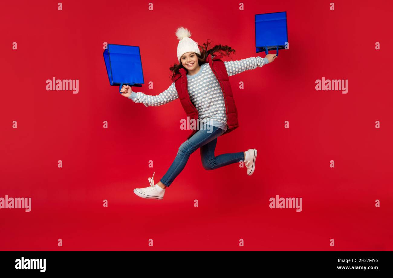 happy teen girl wearing knitted hat jumping with shopping bag on red ...