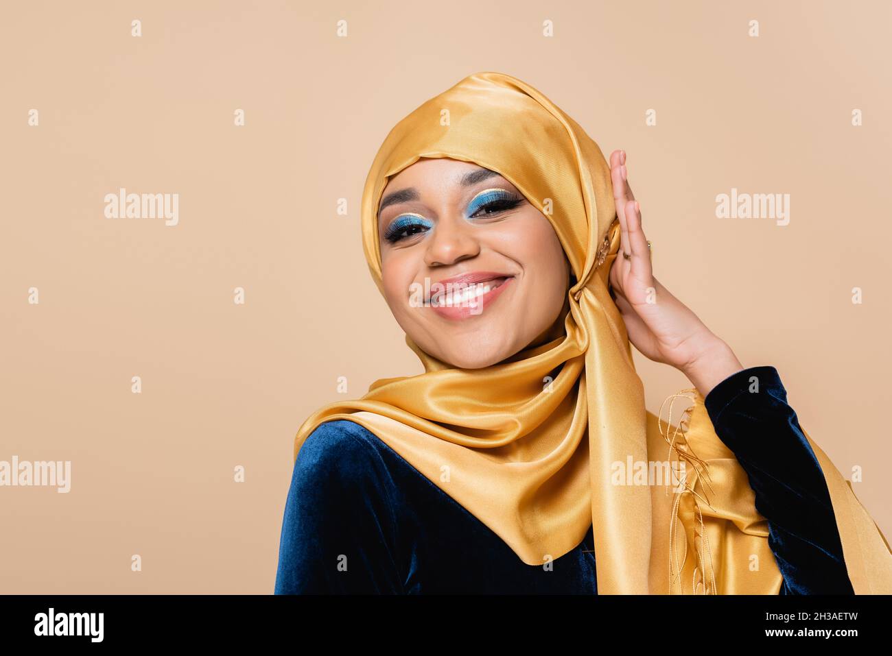 cheerful muslim woman adjusting yellow hijab isolated on beige Stock ...