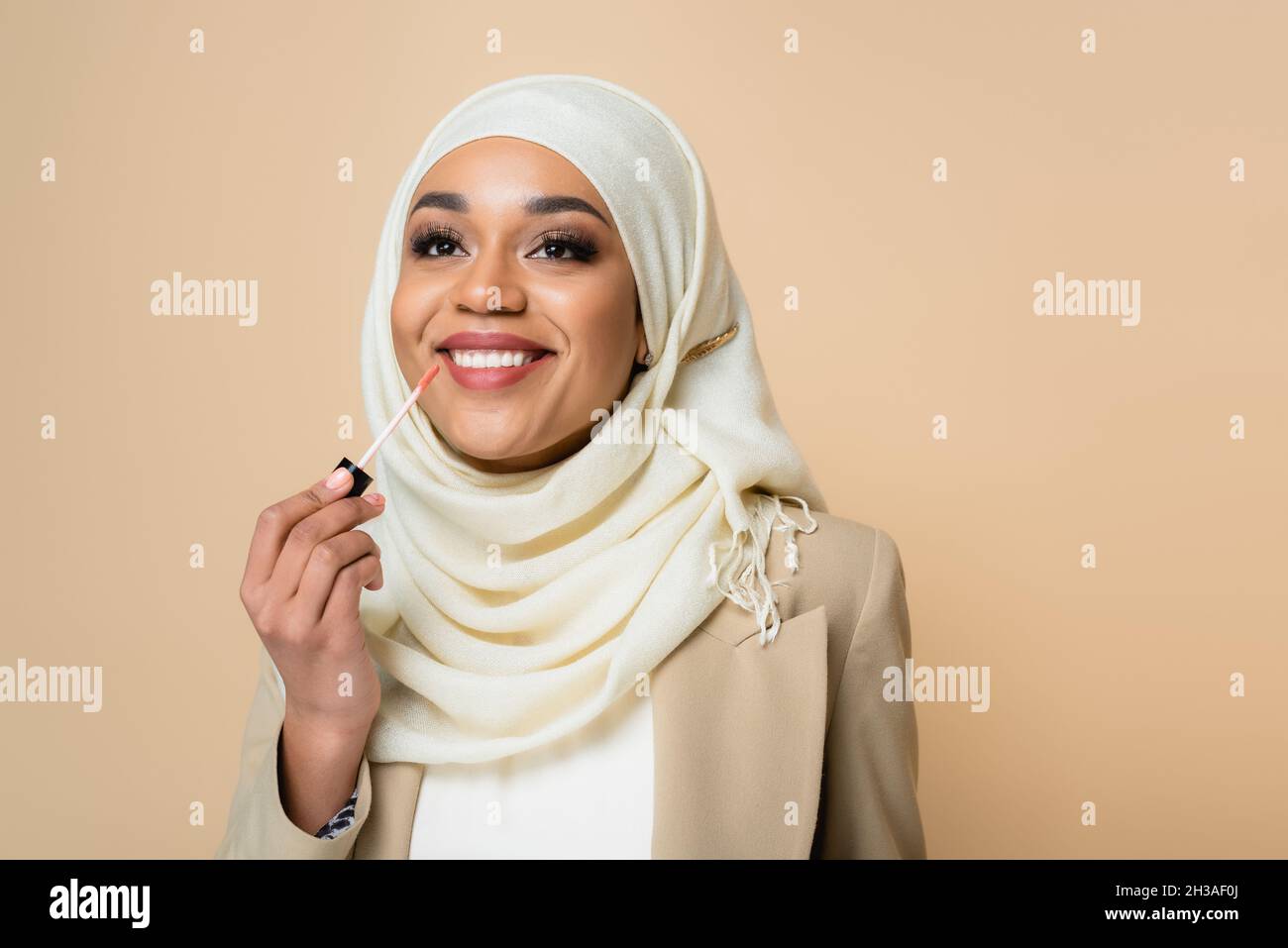 happy muslim woman in hijab applying lip gloss isolated on beige Stock ...