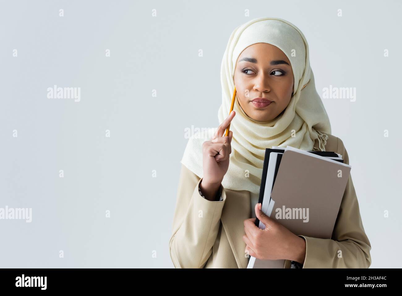 pensive muslim woman in hijab holding pencil and folders isolated on ...