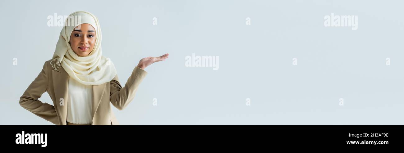 smiling muslim woman in hijab and beige suit pointing with hand ...