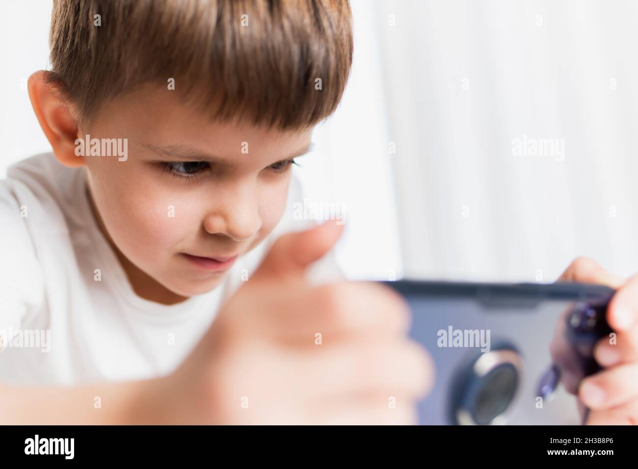 A little boy in a white T-shirt plays games on the phone at home. A ...