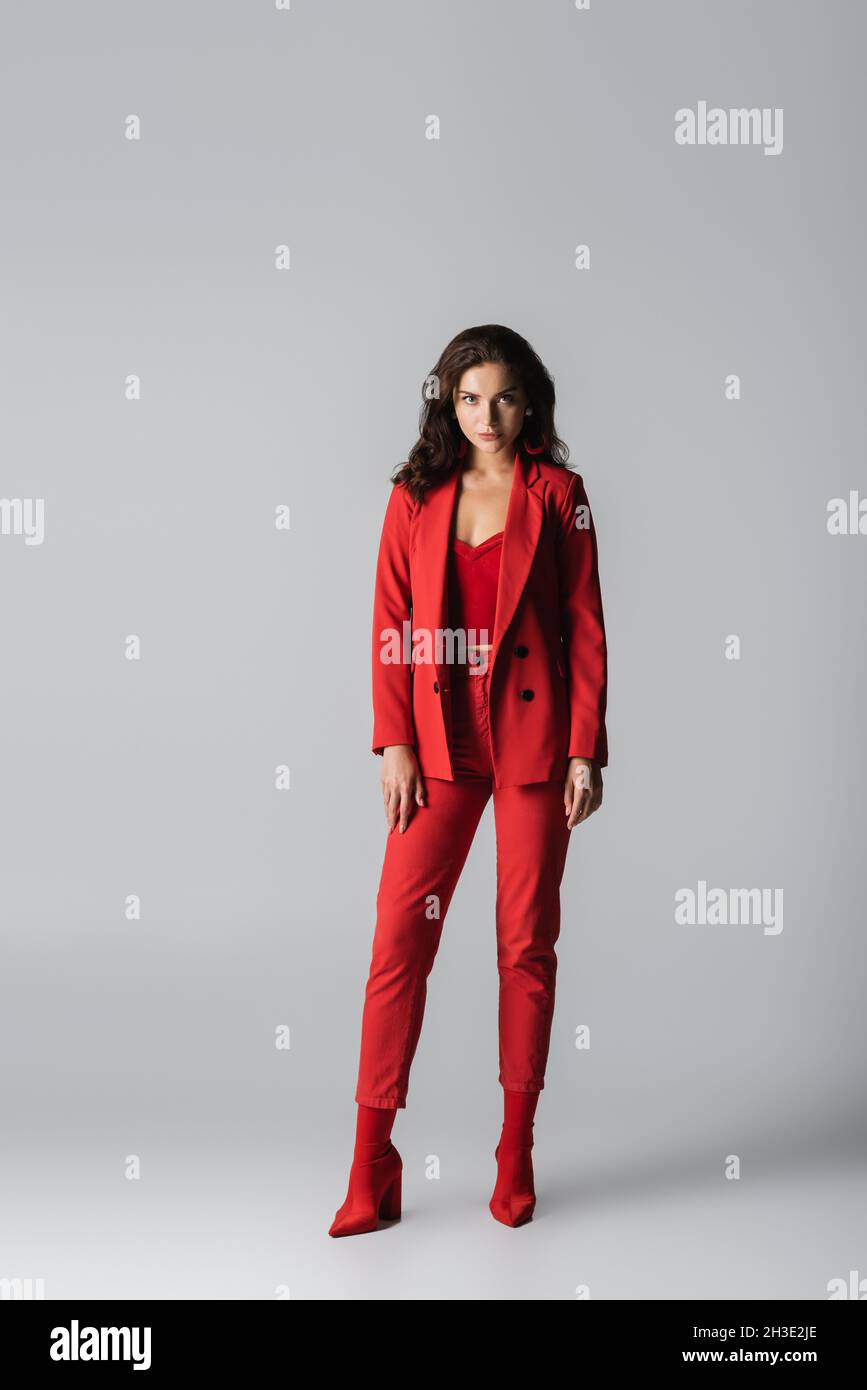 full length of young stylish woman in red boots and suit posing on grey ...