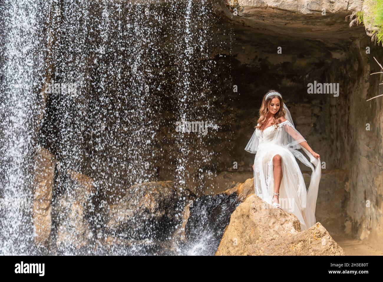 Bride in white wedding dress with veil standing on boulder near falling ...