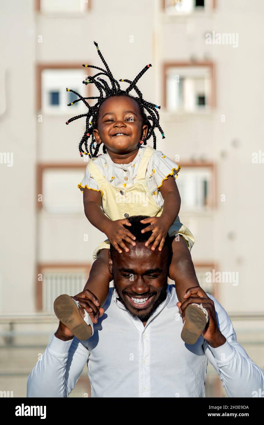Cheerful African American father in shirt carrying little daughter on shoulders and jumping ...
