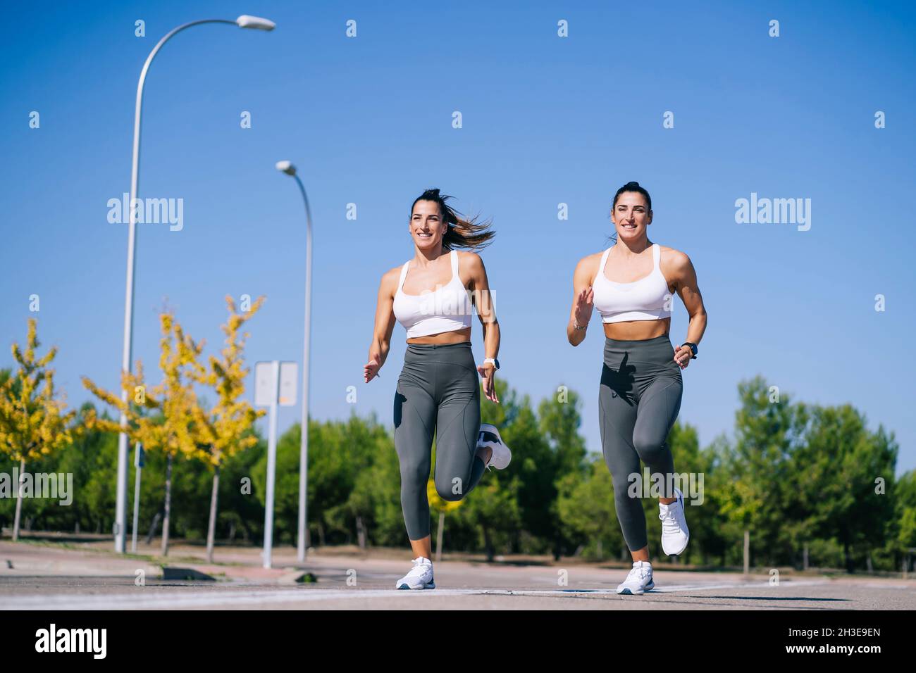 Full body of smiling sporty female twins in sportswear running together ...